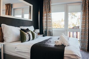 Cama bem arrumada com roupa de cama branca e preta, cabeceira preta e uma janela com cortinas estampadas.