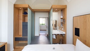 Quarto de hotel moderno com cabeceira de madeira, cama grande, roupeiro embutido, banheiro com pia e espelho, e vista através de uma porta aberta.