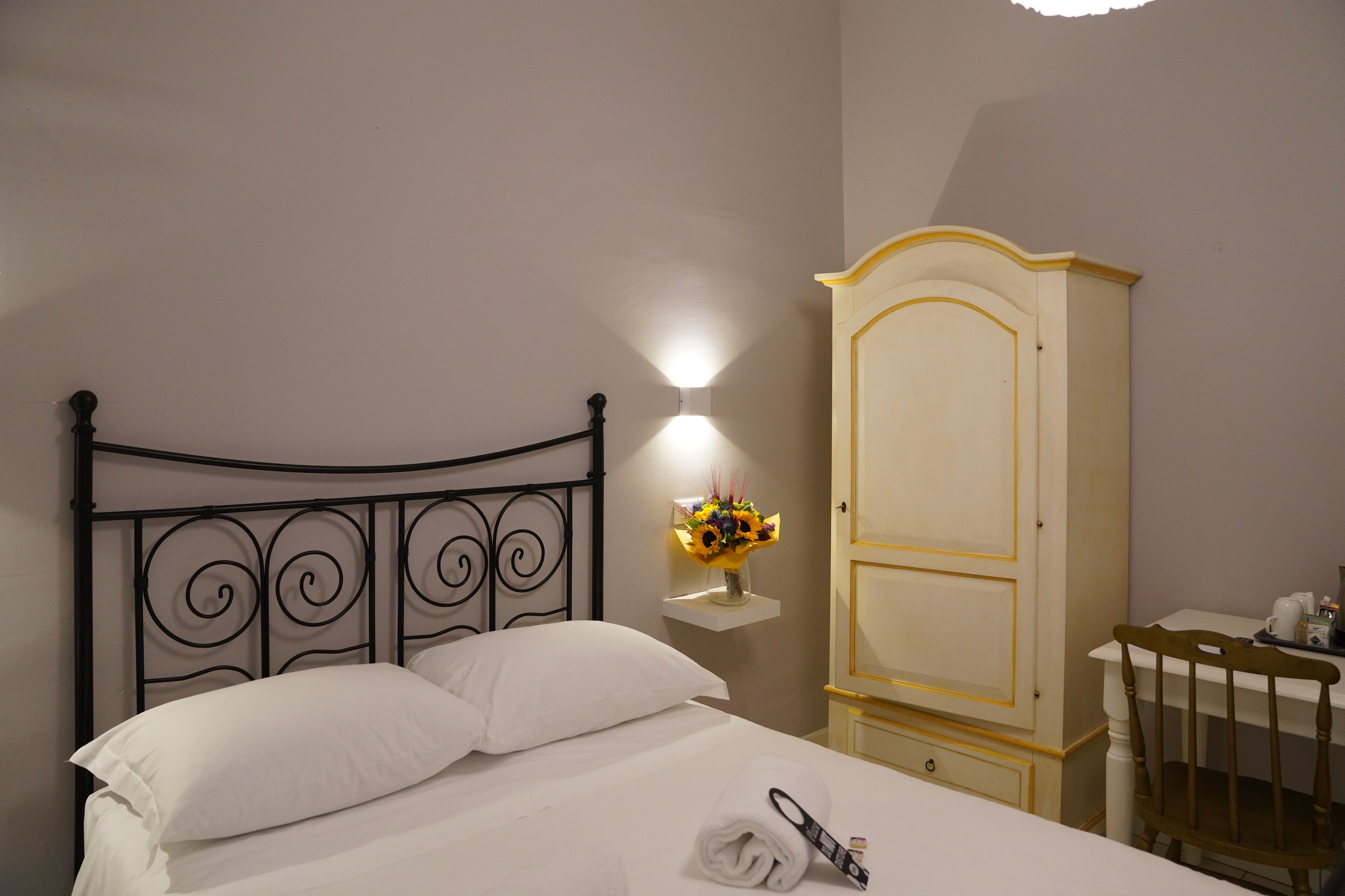 Uma cama bem arrumada com lençóis brancos, uma cabeceira de metal preto, uma mesa de cabeceira com abajur e uma cadeira de madeira.