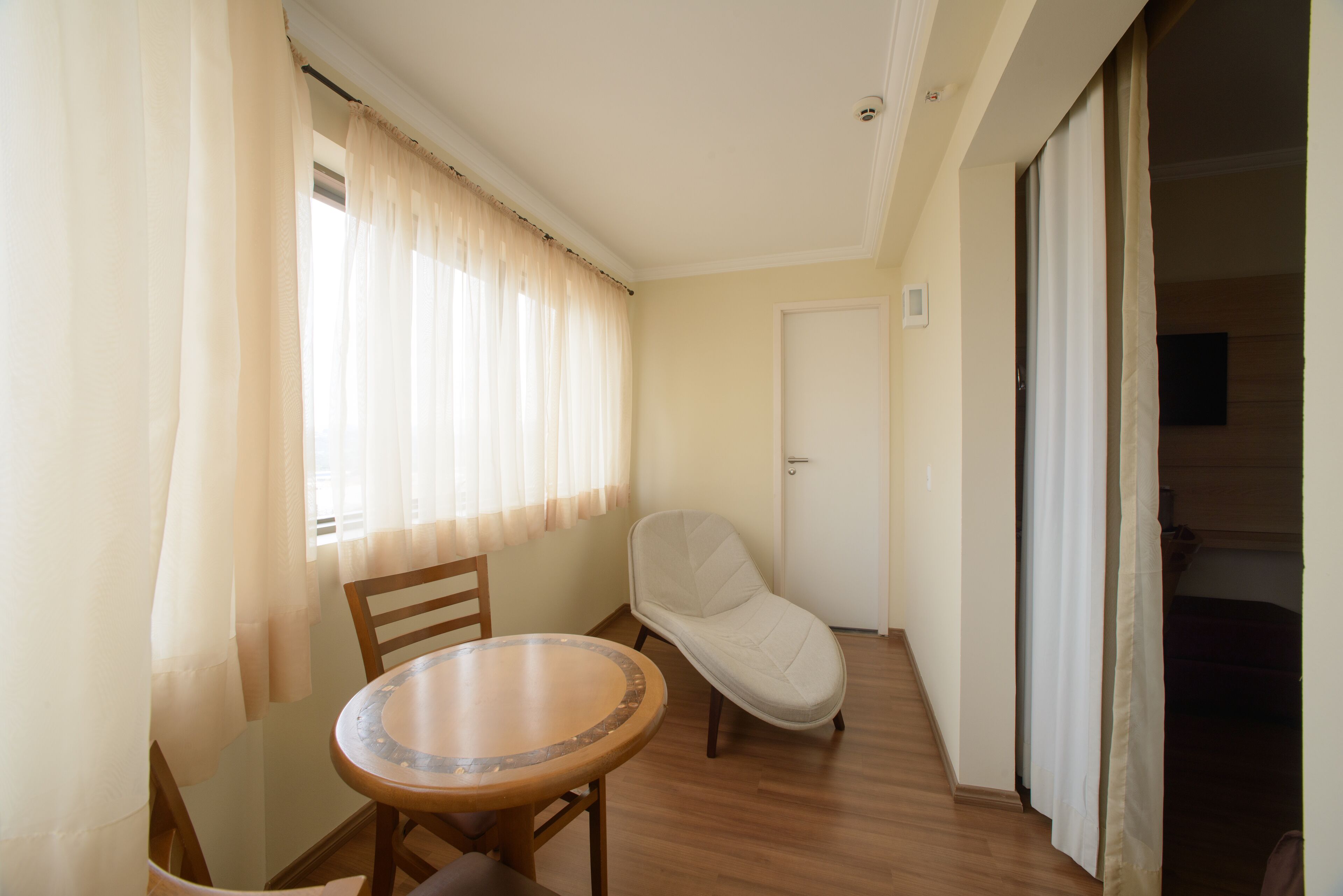 Um quarto com piso de madeira, uma mesa redonda com cadeiras, uma poltrona branca e uma porta.