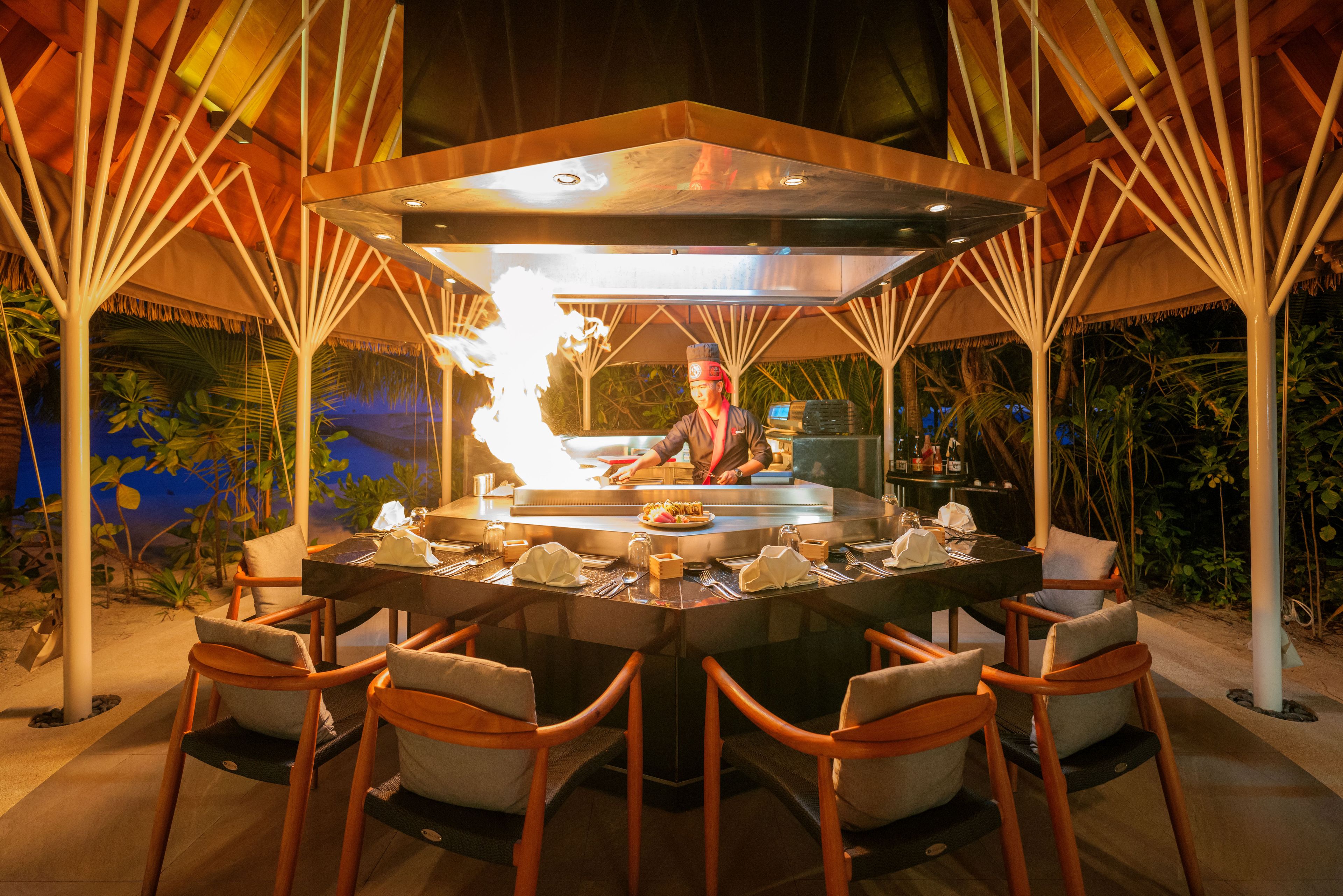 Restaurante com área de cozinha central onde um artista realiza um show com fogo, cercado por mesas e cadeiras.