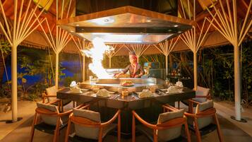 Restaurante com área de cozinha central onde um artista realiza um show com fogo, cercado por mesas e cadeiras.