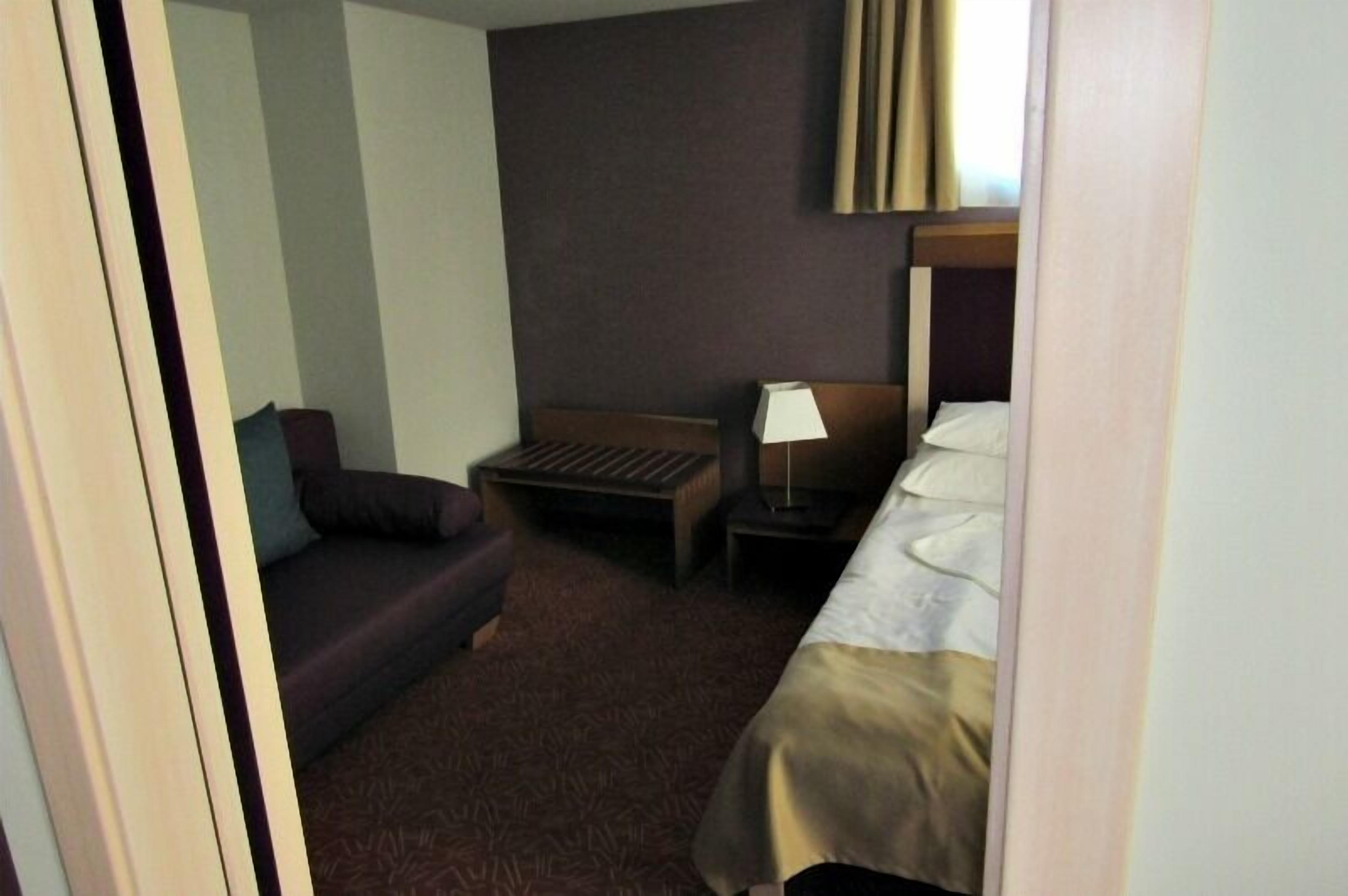 Quarto de hotel com cama, pequena escrivaninha, poltrona, sofá e luminária.