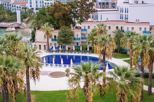 Área de piscina com espreguiçadeiras e guarda-sóis, cercada por palmeiras e um edifício com varandas.