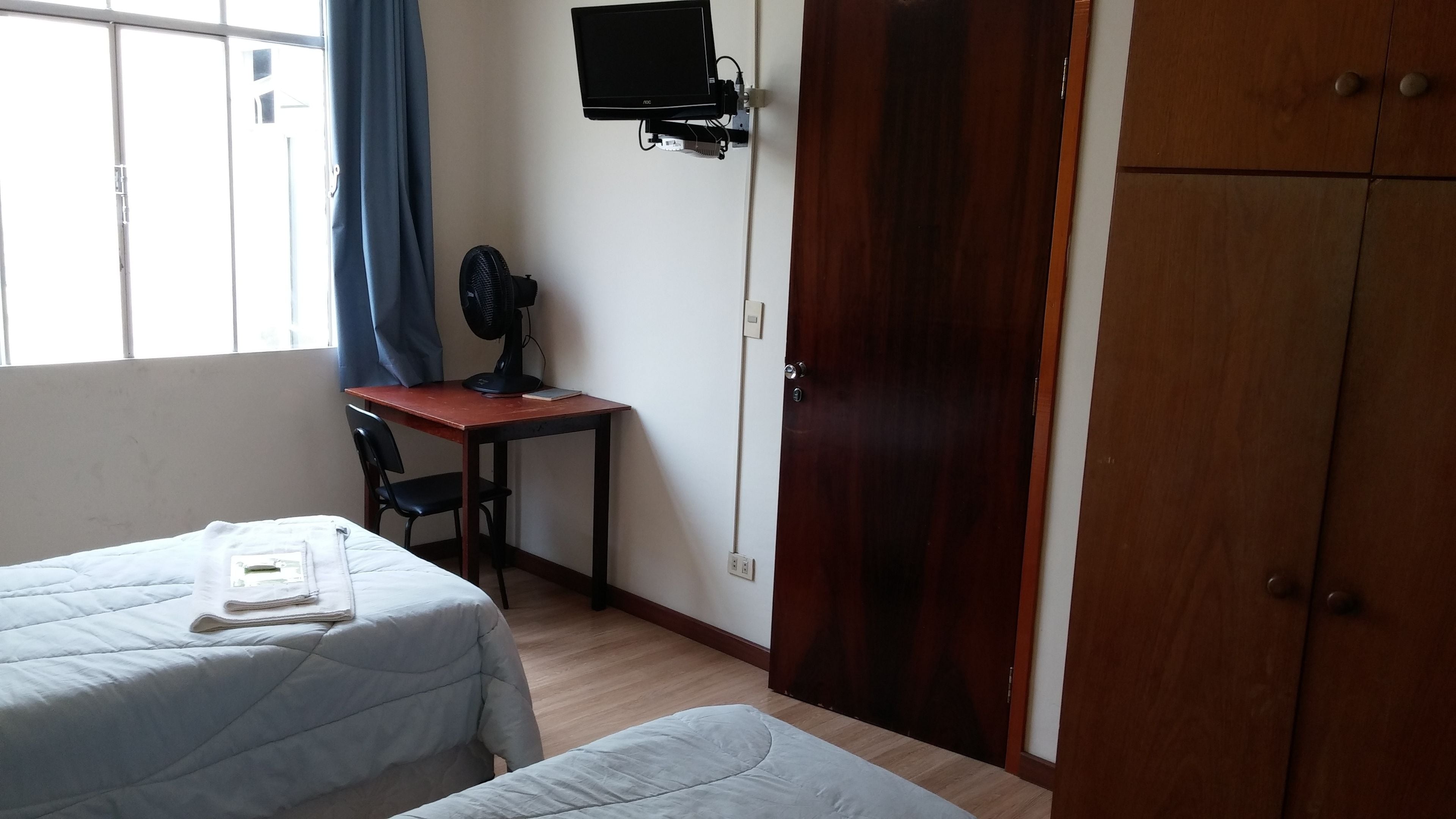 Quarto com duas camas, uma escrivaninha com monitor de computador, um ventilador e um guarda-roupa.