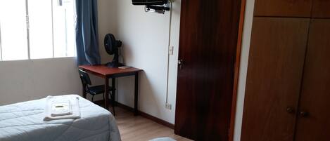 Quarto com duas camas, uma escrivaninha com monitor de computador, um ventilador e um guarda-roupa.