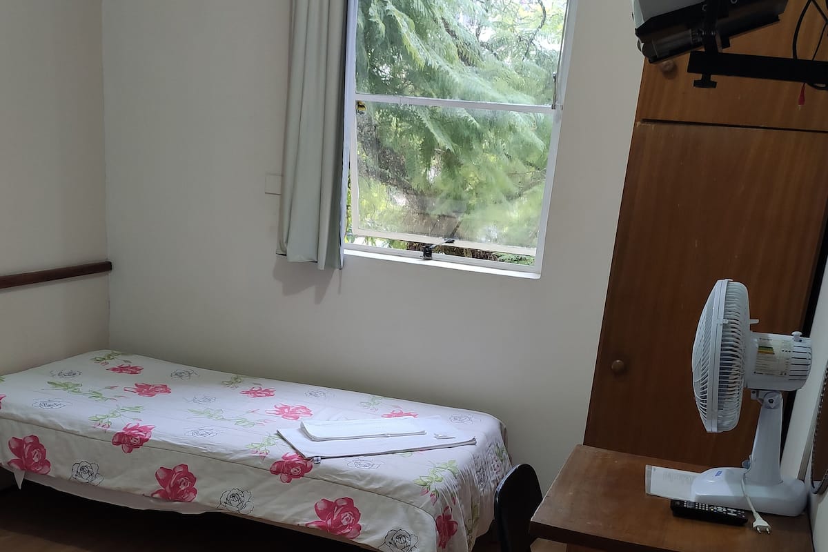 Quarto com duas camas de solteiro, uma escrivaninha com um ventilador, televisão e janela com vista para área verde.