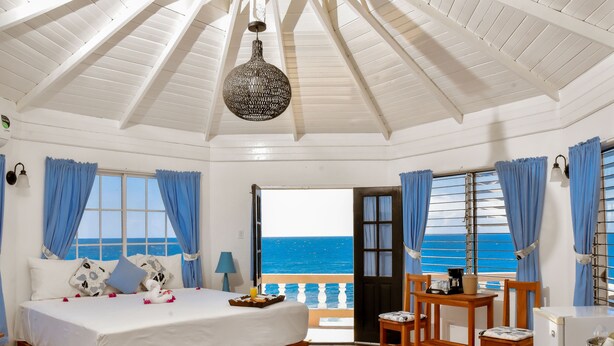 Quarto com cama de madeira, uma mesinha, uma cadeira e vista para o mar pelas janelas.