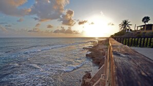 Um calçadão de madeira ao longo da costa, com ondas quebrando na praia e o pôr do sol ao fundo.
