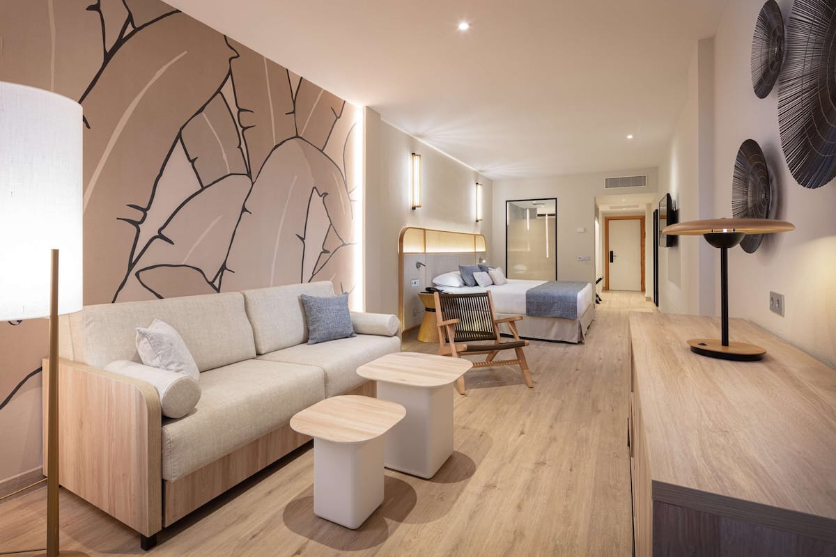 Quarto de hotel moderno com sofá, duas mesinhas laterais, uma cadeira de madeira e cama com cabeceira.