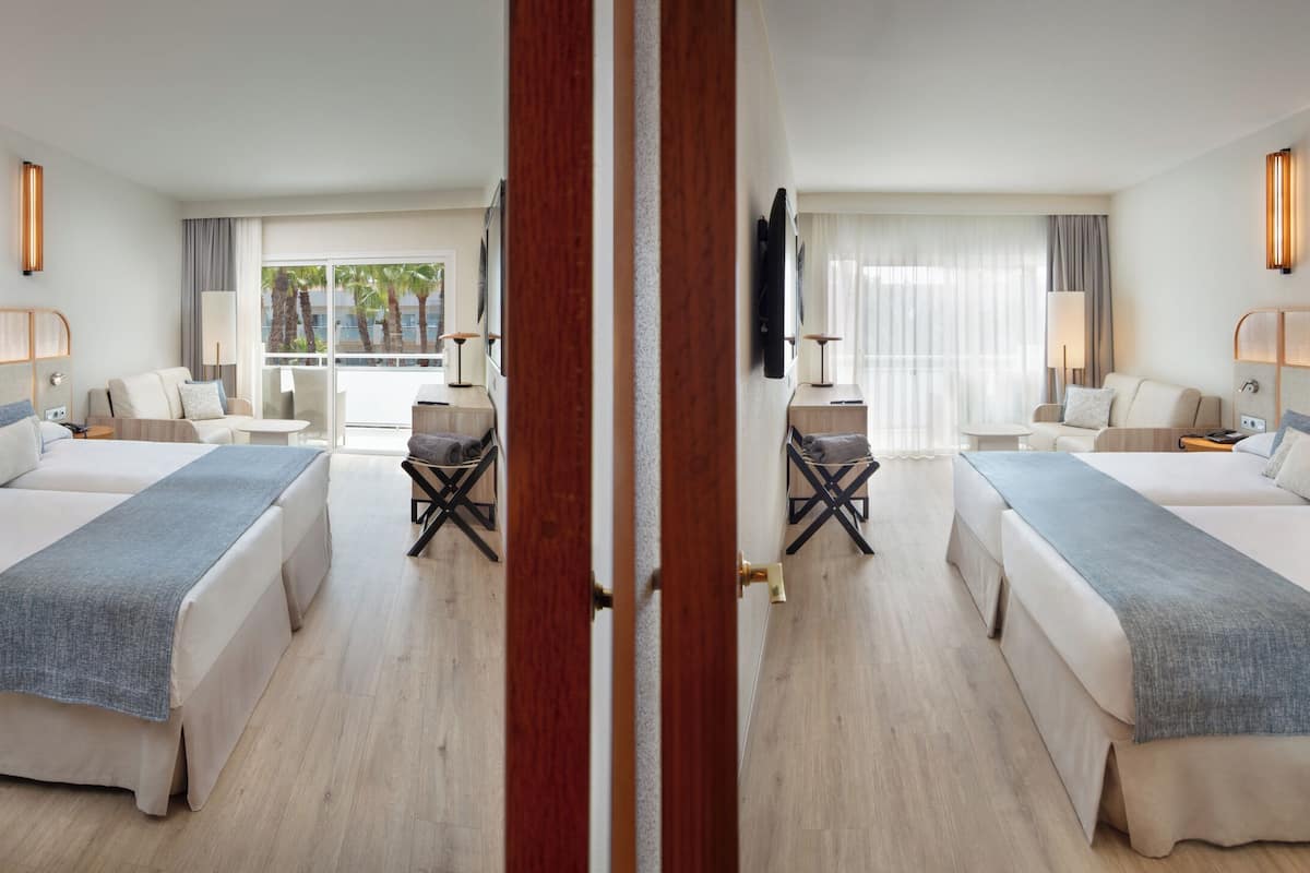 Quarto de hotel com duas camas, um espaço de estar, varanda com vista e uma janela grande.