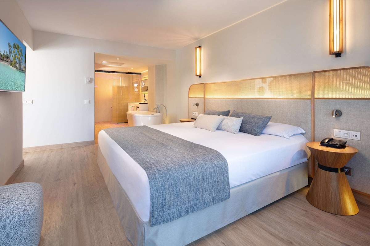 Quarto de hotel moderno com uma cama grande, uma mesinha de cabeceira, banheiro com chuveiro e um quadro na parede.