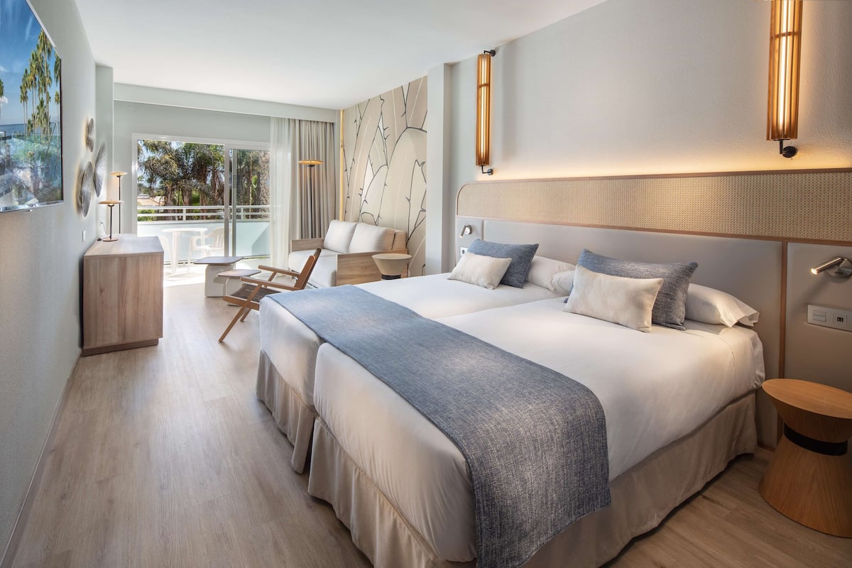 Quarto de hotel moderno com uma cama grande, área de estar com uma cadeira e mesa, e varanda com vista para palmeiras.
