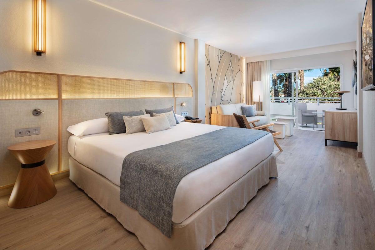 Quarto de hotel moderno com uma cama grande, mesas de cabeceira, escrivaninha e varanda com vista para palmeiras.