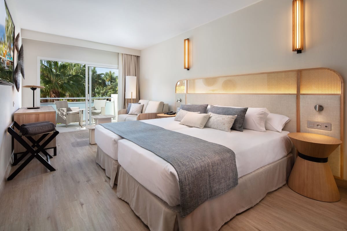 Quarto de hotel moderno com uma cama grande, duas mesas de cabeceira, uma escrivaninha com cadeira e uma varanda com vista para palmeiras.
