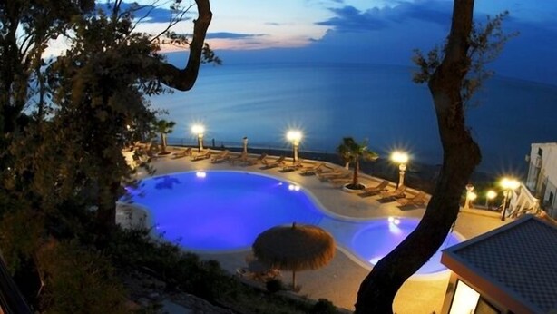 Área de piscina com espreguiçadeiras e um guarda-sol de palha, cercada por árvores e com vista para um corpo d'água.