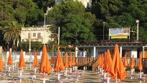 Uma praia com fileiras de espreguiçadeiras e guarda-sóis laranja, uma placa com a inscrição 'Malibu' e um edifício ao fundo.