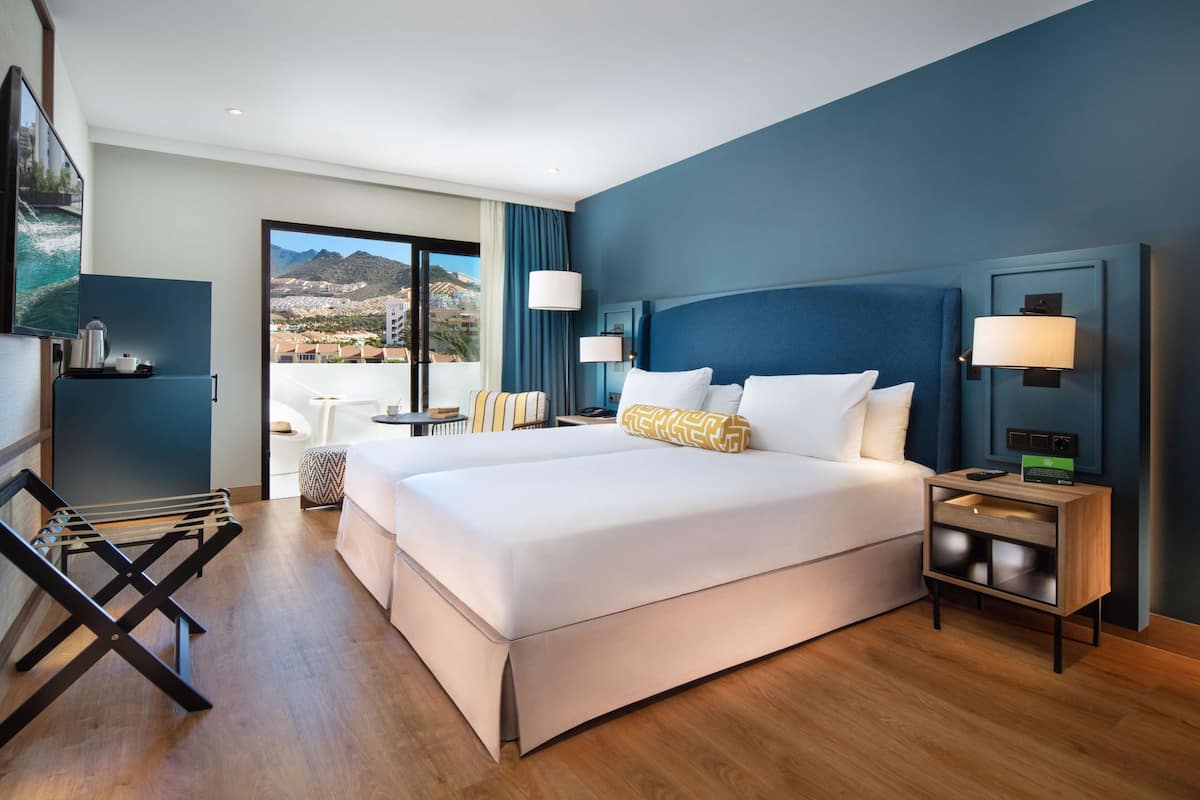 Quarto de hotel moderno com uma cama grande, mesa de cabeceira com luminária, vista para a cidade e varanda.