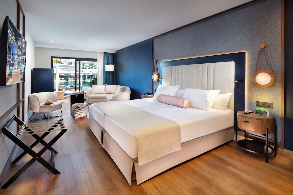 Quarto de hotel moderno com uma cama grande, área de estar com uma poltrona e uma pequena mesa, mesa lateral com luminária e vista para a área da piscina pela janela.