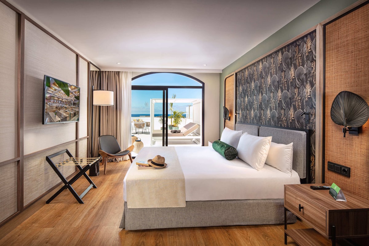 Quarto de hotel moderno com uma cama grande, vista para o mar, área de estar com mesa e cadeiras, e uma grande obra de arte na parede.
