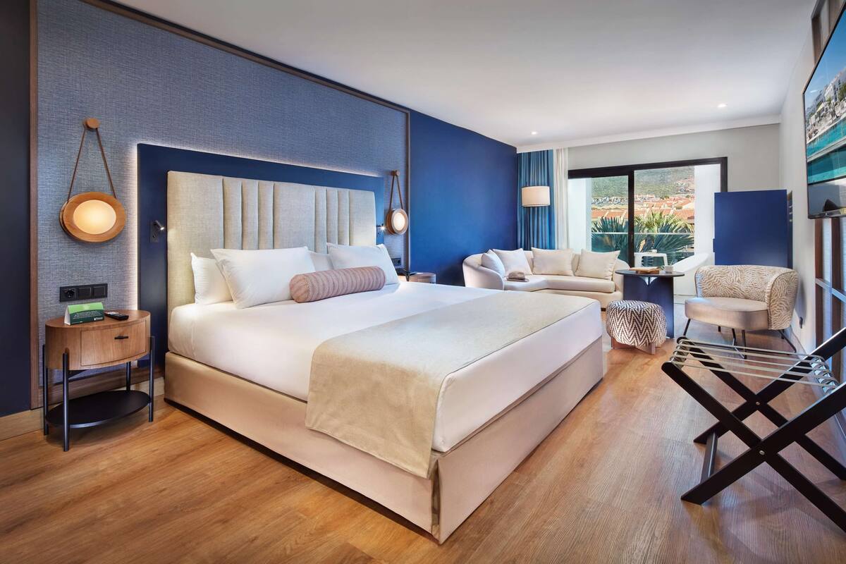 Quarto de hotel moderno com uma cama grande, um sofá, uma poltrona e uma televisão.