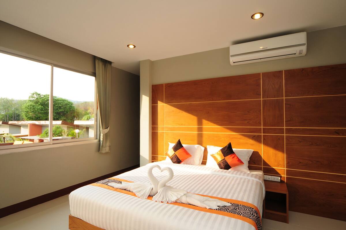 Quarto de hotel com uma cama grande, cabeceira de madeira e janela com vista para vegetação.