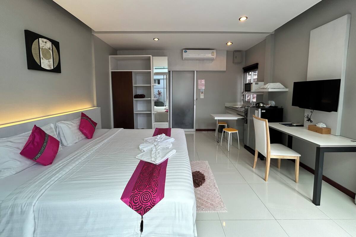 Quarto de hotel com uma cama grande, uma escrivaninha, uma cadeira, uma TV e uma cozinha compacta.