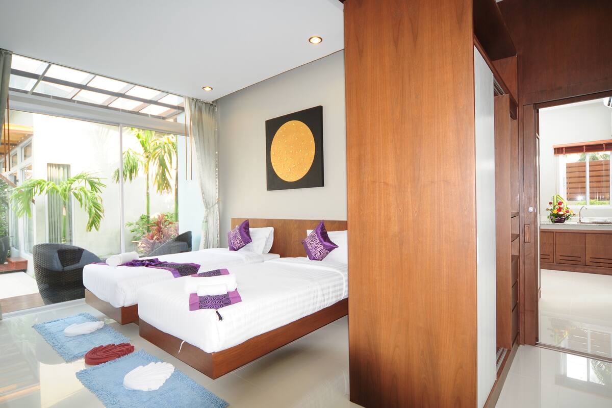 Quarto de hotel moderno com uma cama grande, cabeceira de madeira e vista para palmeiras através de amplas janelas.