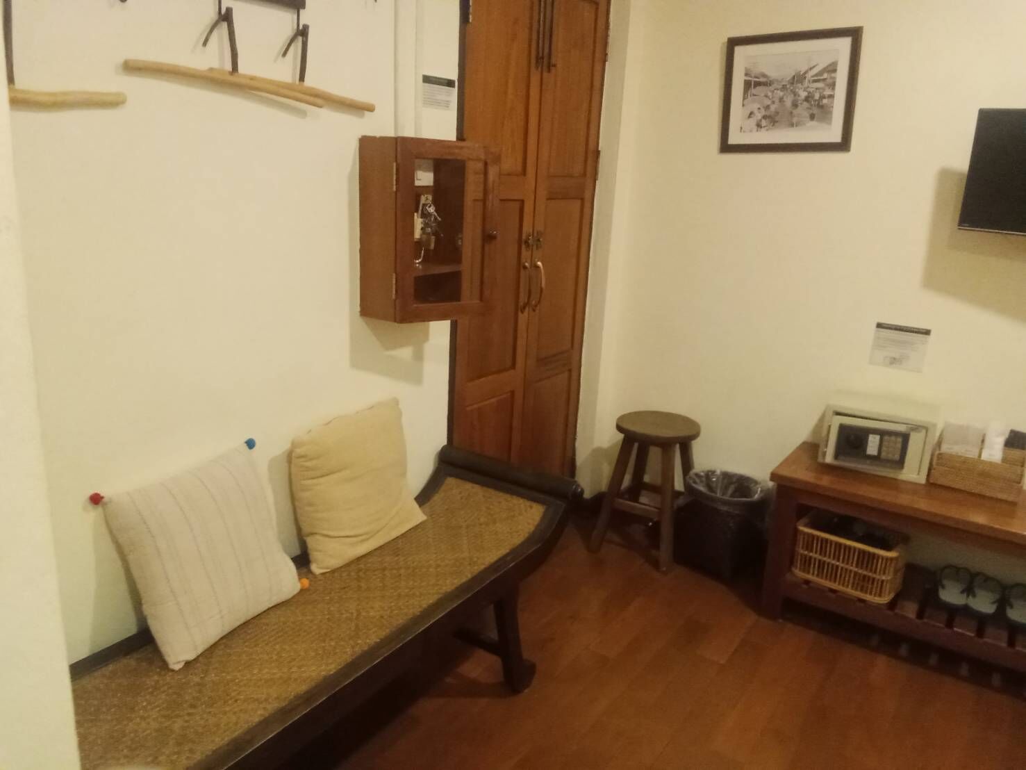Um quarto com um banco de madeira, um banquinho, uma lixeira e uma estante com garrafas.