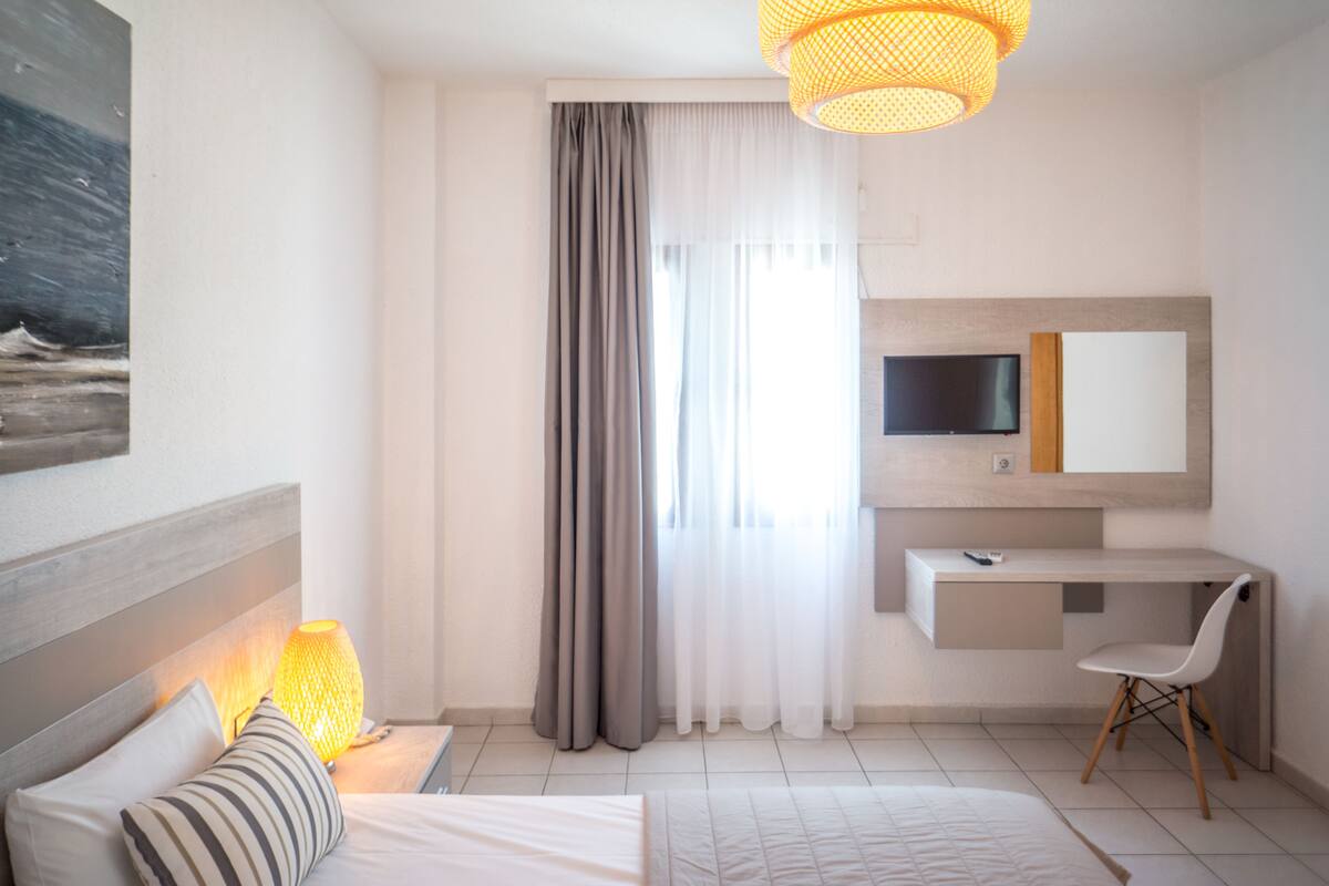 Quarto de hotel moderno com cama, abajur, escrivaninha, cadeira, televisão e uma janela grande com cortinas.