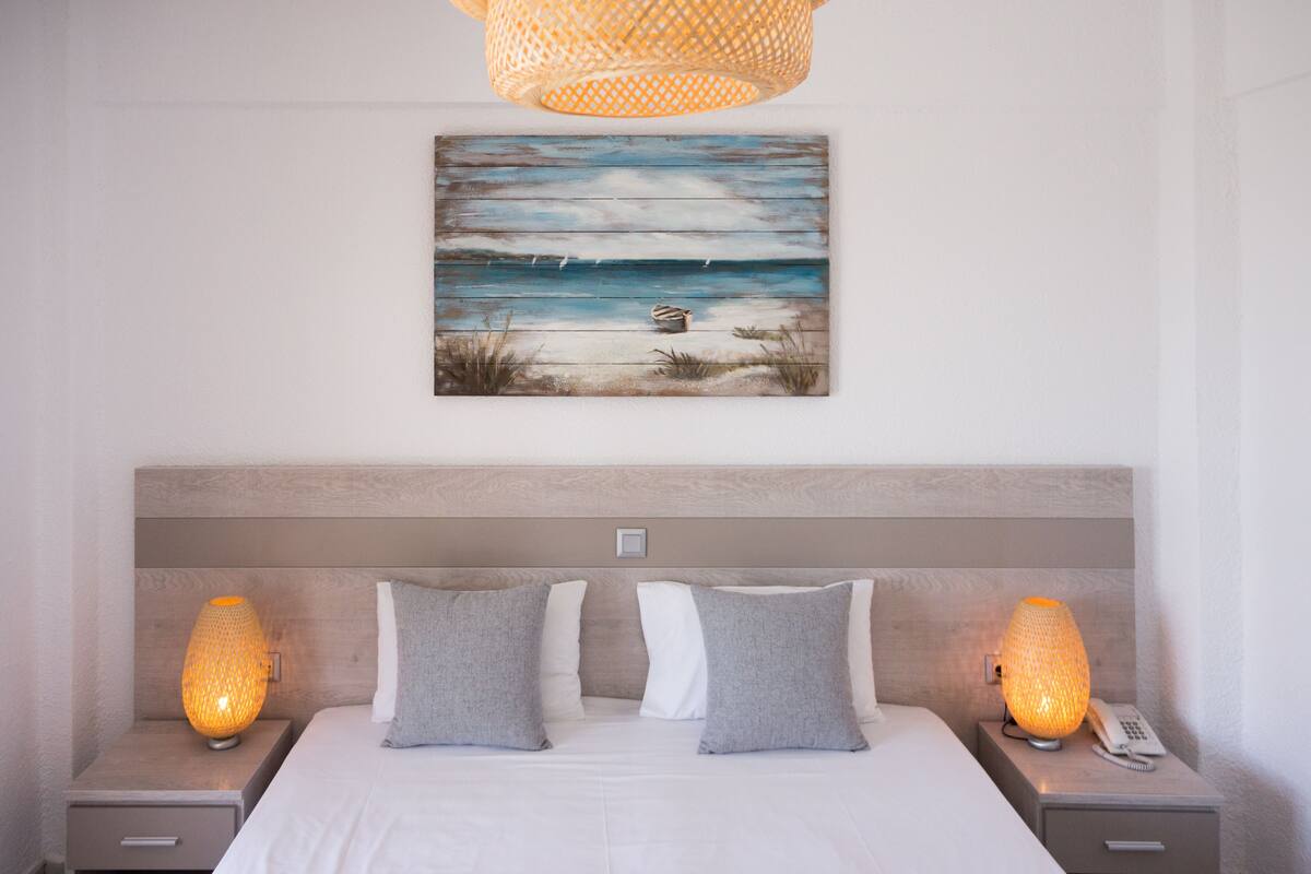 Quarto com cama, duas luminárias de cabeceira, pintura de uma praia e cabeceira de madeira.
