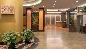 Um saguão de hotel com uma mesa de recepção, plantas em vasos, um painel decorativo e um ventilador de teto.