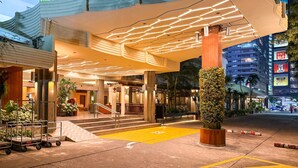 Entrada de hotel com design moderno, incluindo uma área coberta com iluminação geométrica, uma escadaria que leva ao saguão, e um aviso da KFC visível ao fundo.