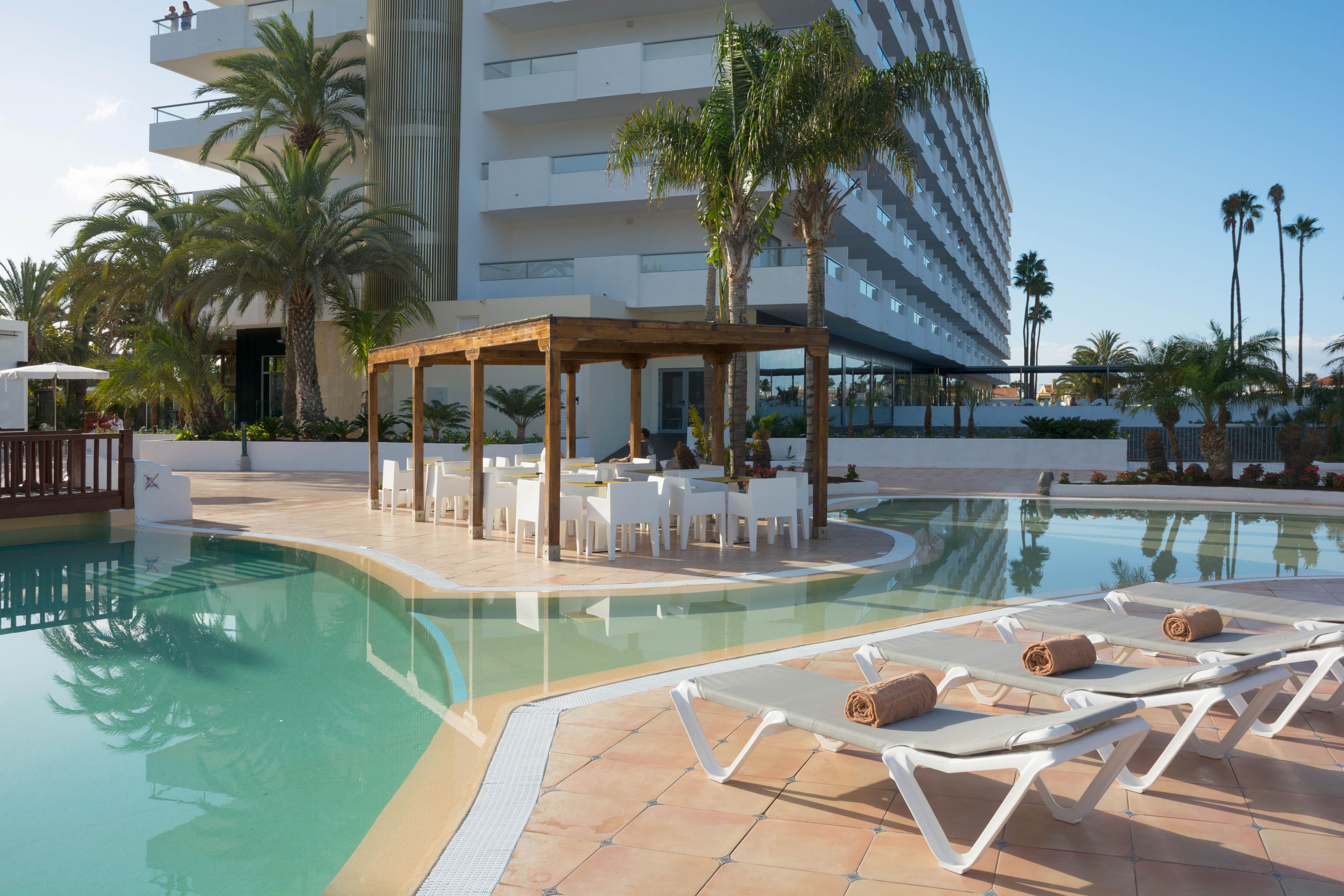 Uma área de piscina com espreguiçadeiras, uma pérgula de madeira e palmeiras.