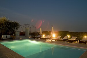 Área de piscina com espreguiçadeiras e guarda-sóis, cercada por um jardim ajardinado e vista para o horizonte da cidade ao entardecer.