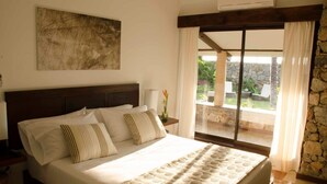 Cama bem arrumada com lençóis brancos e almofadas listradas, uma grande pintura abstrata na parede e vista para uma parede de pedra e vegetação através de uma porta de vidro.