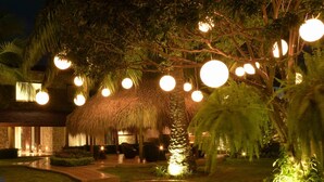 Área externa bem iluminada com estrutura de telhado de palha, cercada por vegetação exuberante e iluminada por esferas de luz.