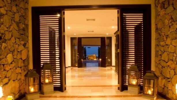 Entrada de hotel com paredes de pedra e chão quadriculado, conduzindo a um quarto com cama e vista para o céu.