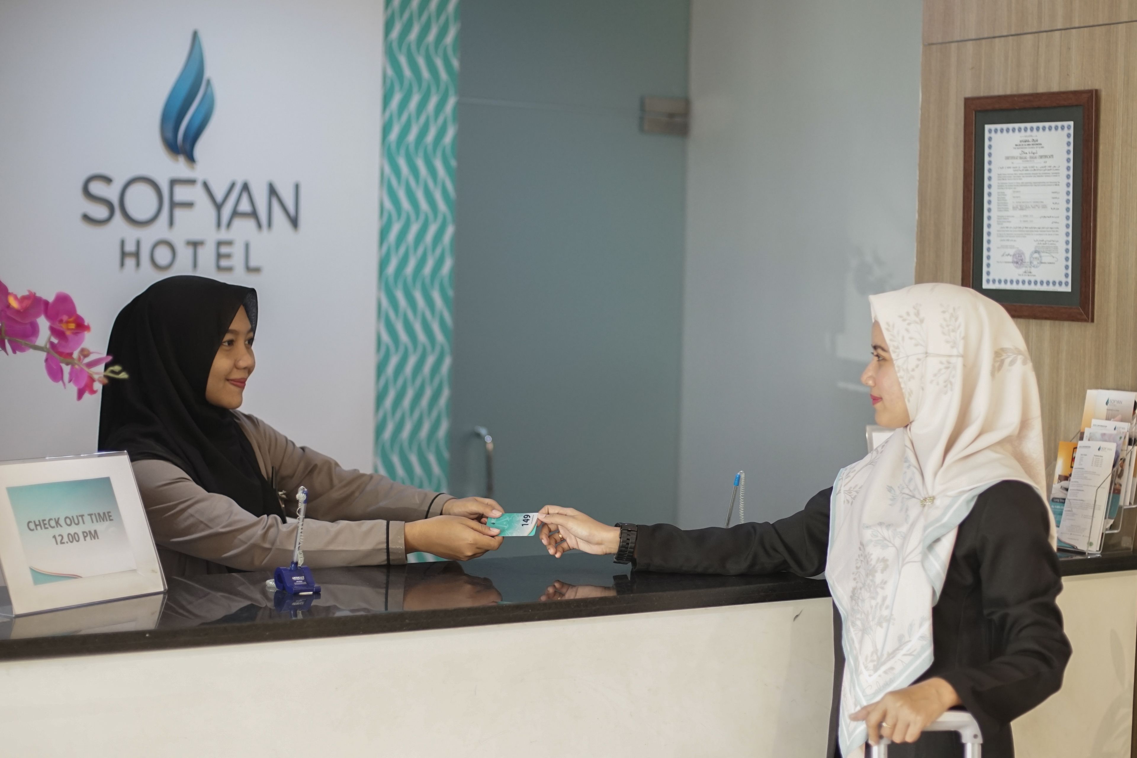 Bancada de recepção do Sofyan Hotel, com indicação do horário de check-out, um certificado emoldurado e uma parede decorativa com um logotipo azul.