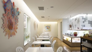 Um restaurante moderno com mesas e cadeiras brancas, uma área de cozinha e uma pintura abstrata colorida na parede.