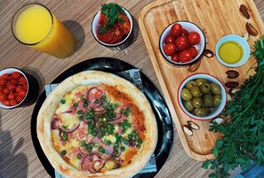 Uma pizza com diversos ingredientes em um prato preto, cercada por potes de tomate, azeitonas e um copo de suco de laranja em uma mesa de madeira.
