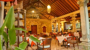 Um restaurante com mesas e cadeiras de madeira, piso de azulejos e teto alto com vigas aparentes.