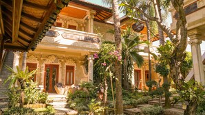 Um edifício tradicional com entrada coberta, escadaria que leva ao andar superior e um jardim com diversas plantas e um bebedouro para pássaros.