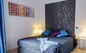 Cama bem arrumada com manta e almofadas azuis, parede com padrão floral e uma pintura abstrata colorida acima da cama.