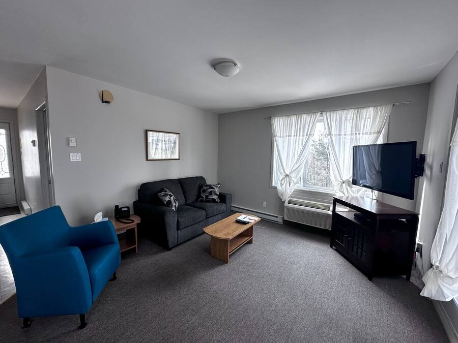 Uma sala de estar moderna com um sofá cinza, uma poltrona azul, uma mesa de centro de madeira, uma televisão de tela plana e uma janela grande com cortinas translúcidas.