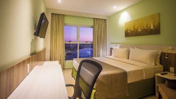 Quarto de hotel com cama, escrivaninha, cadeira, televisão e um quadro na parede.