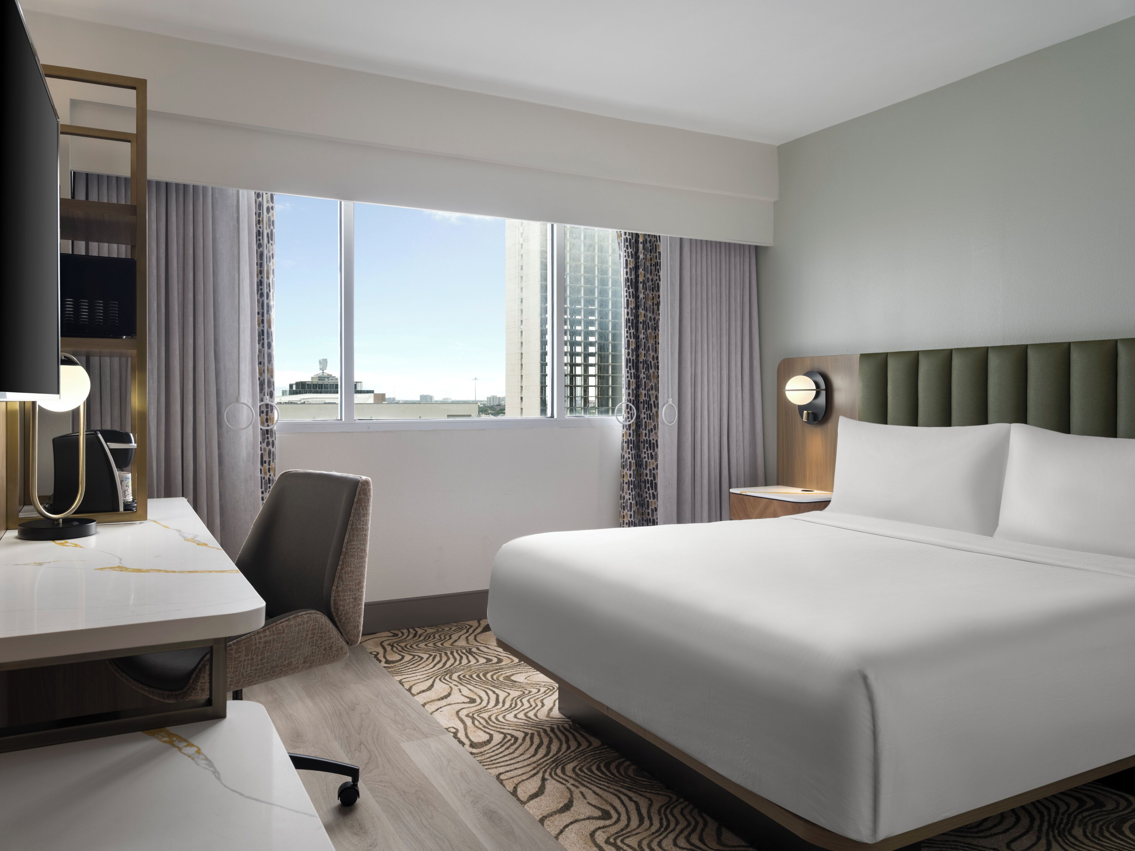 Quarto de hotel com cama, escrivaninha, cadeira e vista para a cidade.