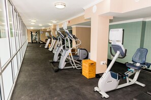 Uma academia com várias esteiras e bicicletas ergométricas, um cesto de madeira e uma televisão fixada na parede.