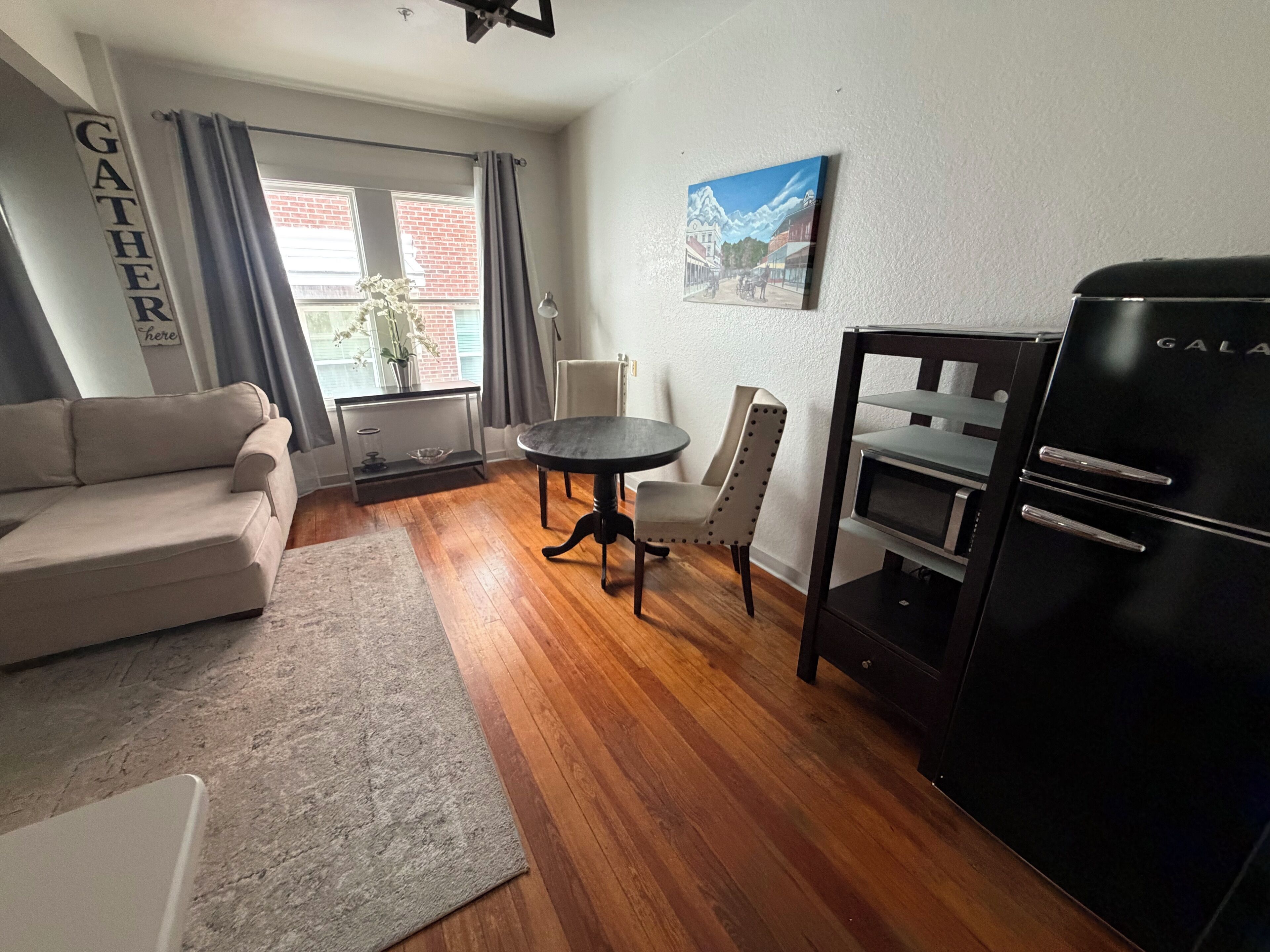Uma sala de estar com sofá, mesa de jantar com cadeiras, geladeira preta e um quadro na parede.