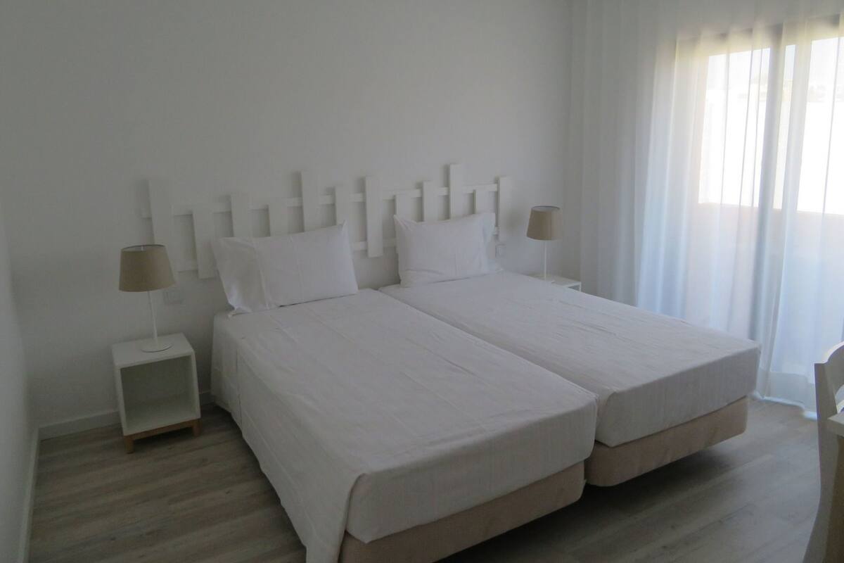 Uma cama de casal com cabeceira branca, duas mesas de cabeceira com luminárias e uma janela grande com cortinas leves.